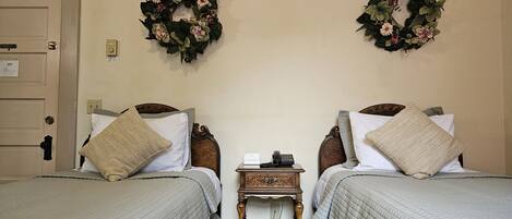 Quarto de hotel com duas camas, cada uma com um travesseiro e um edredom acolchoado. Há duas guirlandas fixadas na parede, uma mesa de cabeceira de madeira com telefone, e uma porta com fechadura e interruptor de luz.