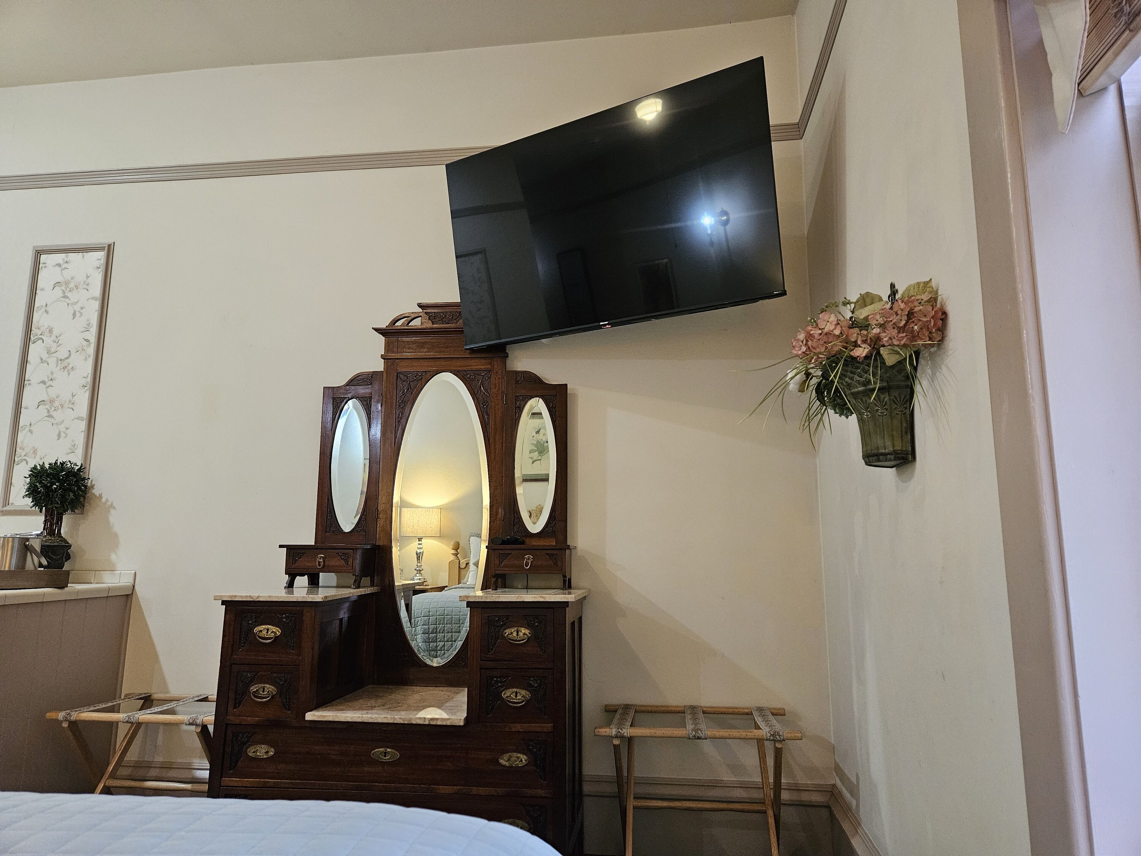 Quarto com cômoda de madeira, televisão de tela plana na parede, cama e pequena mesa com luminária.