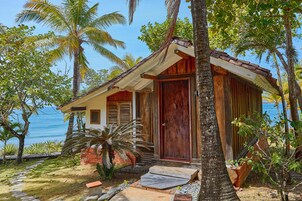 Uma casa de madeira com porta vermelha, cercada por palmeiras e vista clara do oceano.