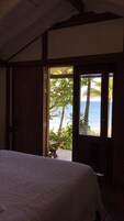 Quarto com cama, porta de madeira e vista para a praia e oceano através de uma janela.