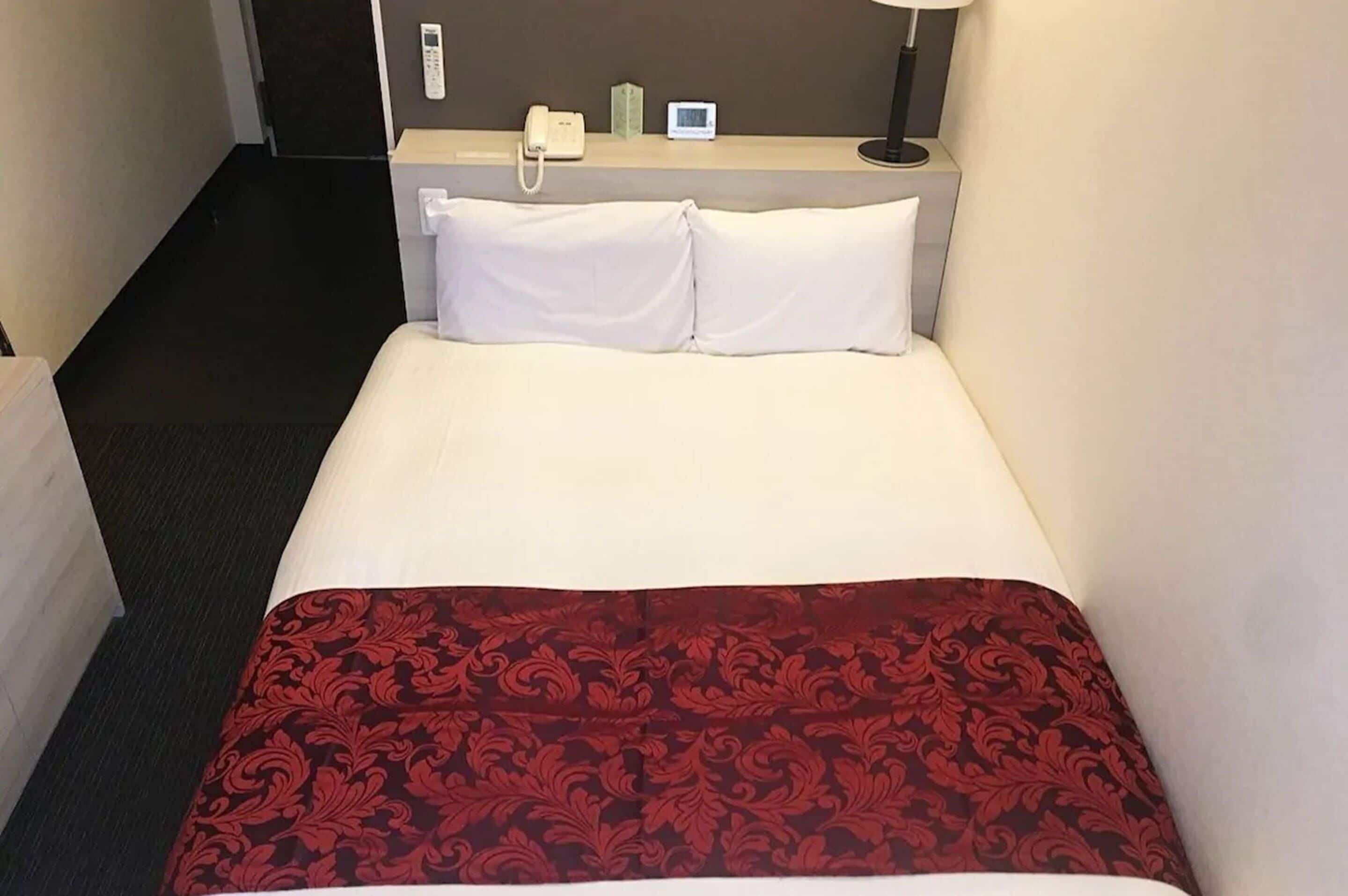 Quarto de hotel com cama, telefone ao lado da cama, relógio digital e abajur na mesa de cabeceira.
