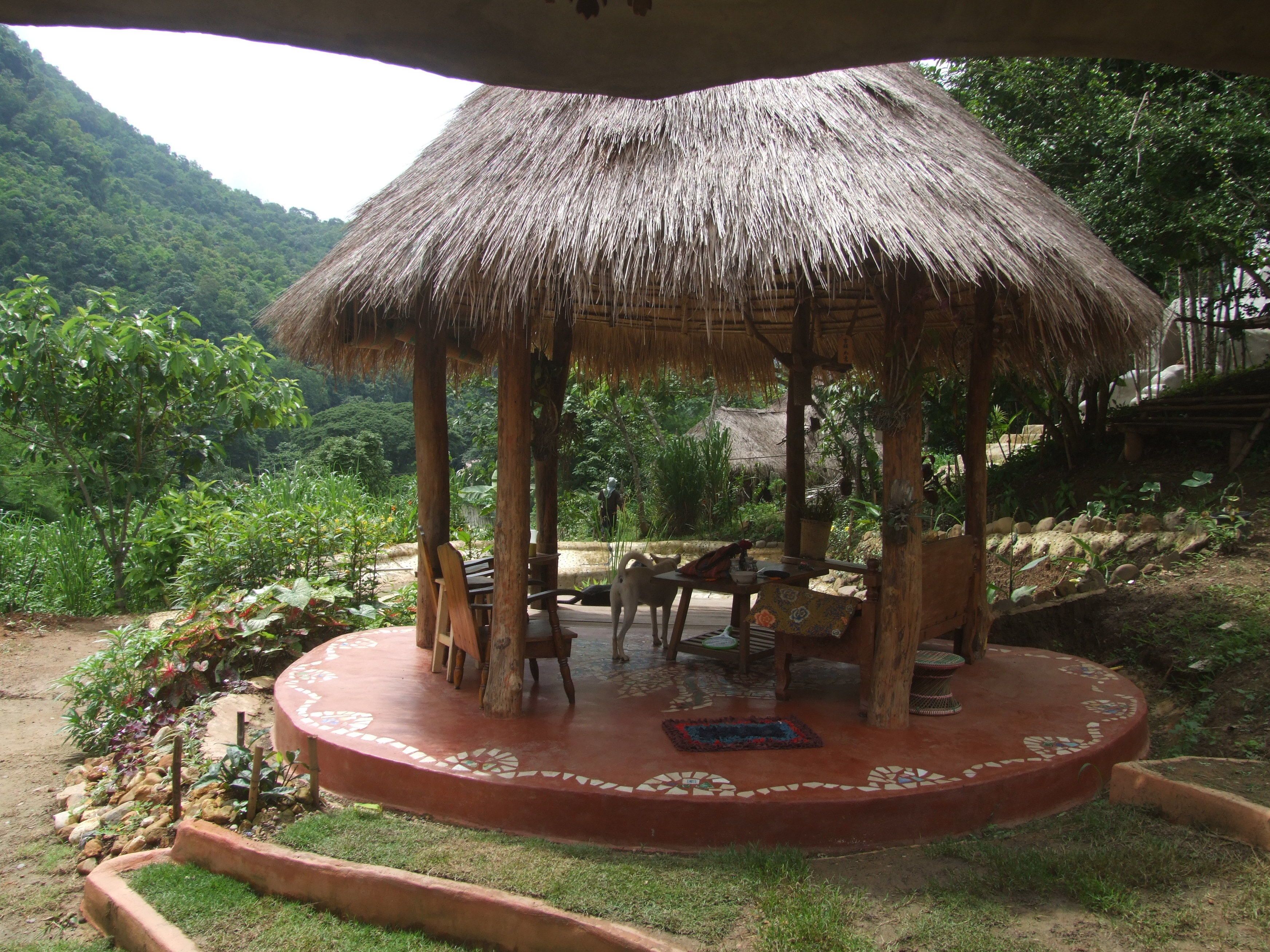 Um gazebo com telhado de palha e piso circular, rodeado por um jardim com diversas plantas e um banco.