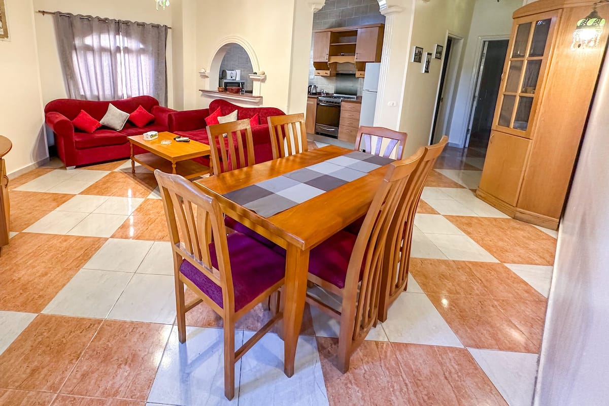 Uma sala de jantar com mesa e cadeiras de madeira, um sofá vermelho e uma cozinha ao fundo.