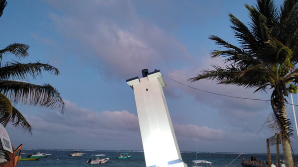 Uma torre branca e alta com uma bandeira no topo, rodeada por palmeiras e barcos na água.