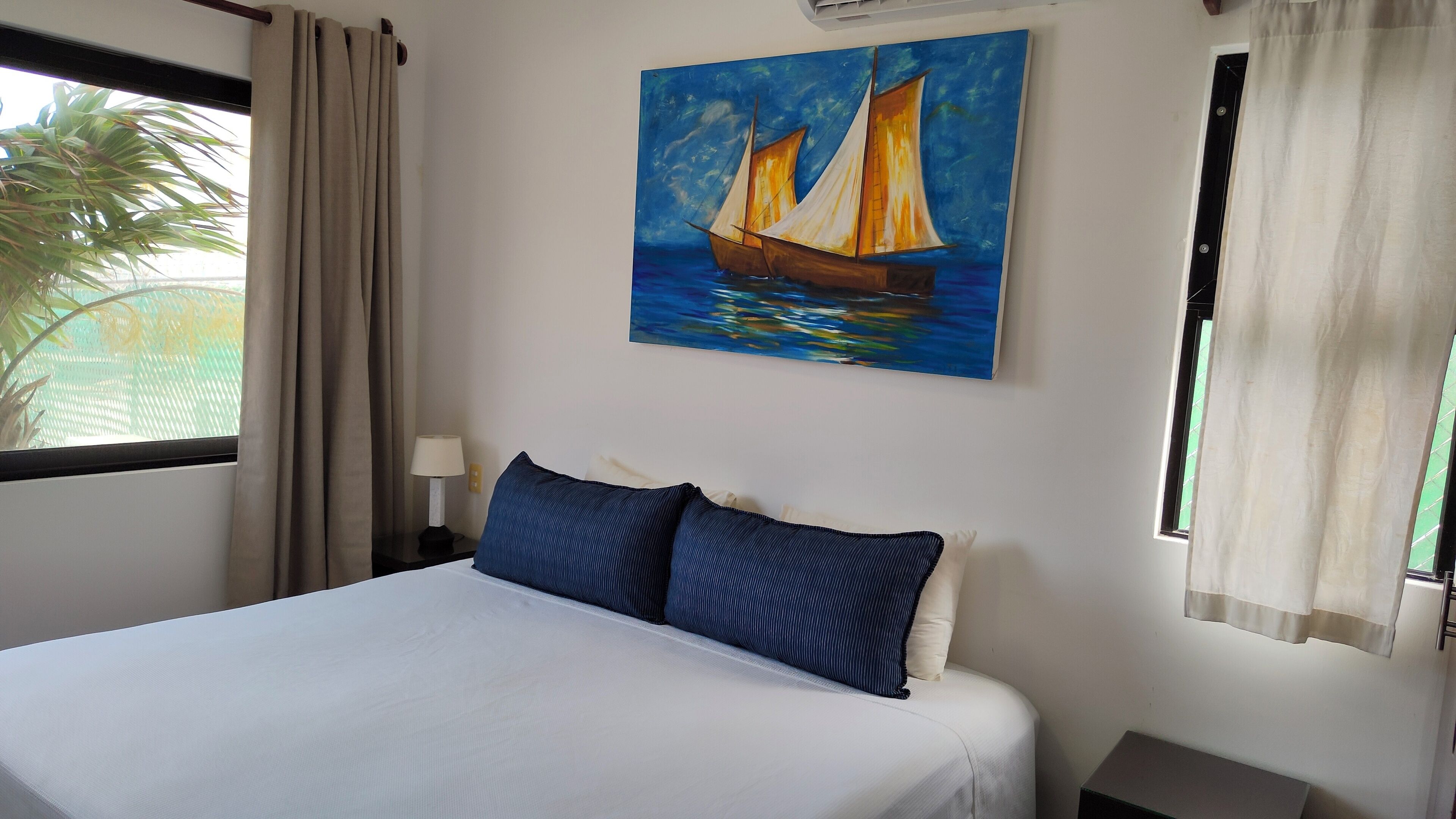 Quarto de hotel com cama, pintura de um veleiro, mesa de cabeceira e janela com cortinas.