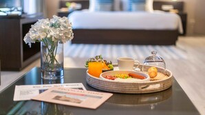 Quarto de hotel com bandeja de café da manhã bem arrumada sobre uma mesa, um vaso com flores e uma cama com almofadas ao fundo.