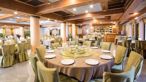 Uma sala de jantar ampla com mesas redondas, cadeiras estofadas em verde e um bar ao fundo.