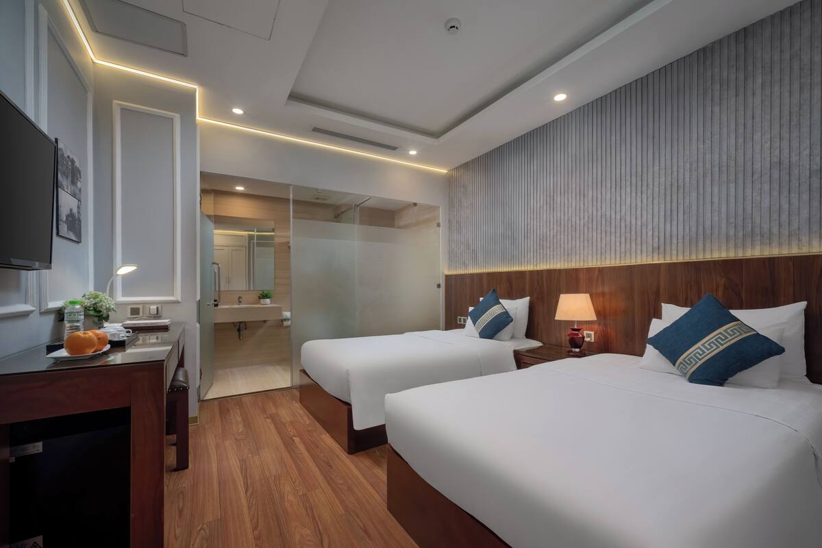 Quarto de hotel com duas camas, uma escrivaninha, uma televisão e um banheiro visível por uma porta de vidro.