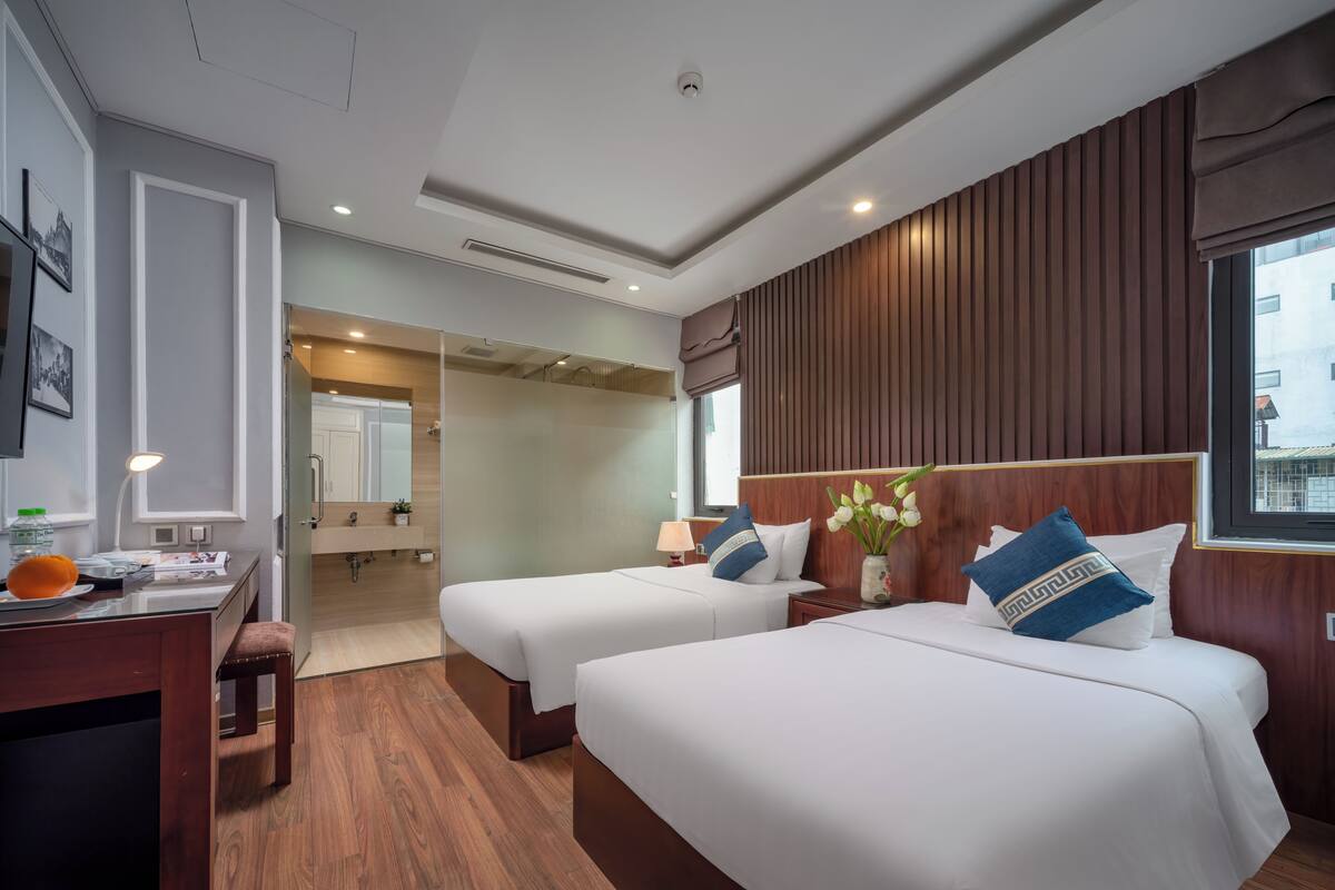 Quarto de hotel com duas camas, uma escrivaninha, um espelho e uma janela com vista.