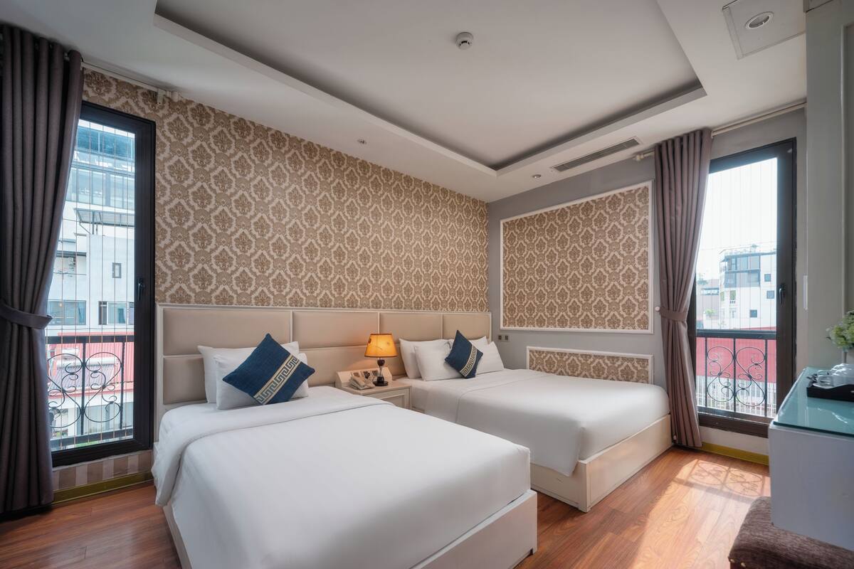 Quarto de hotel com duas camas, uma mesa de cabeceira, uma luminária e vista para edifícios por grandes janelas.
