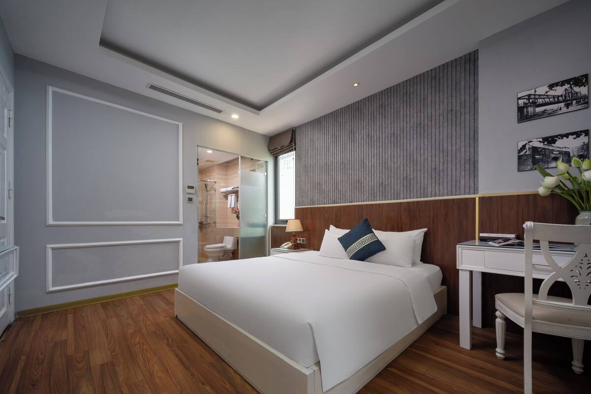 Quarto de hotel moderno com uma cama grande, mesa de cabeceira com abajur, uma cadeira e vista para o banheiro.