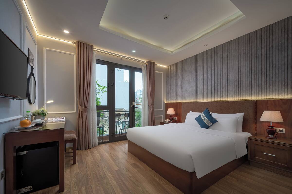 Quarto de hotel com uma cama grande, escrivaninha, televisão, varanda com cortinas e chão de madeira.