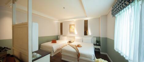 Quarto de hotel com duas camas, uma escrivaninha, uma cadeira, uma mesinha de centro com um vaso de flores e uma janela com cortinas.