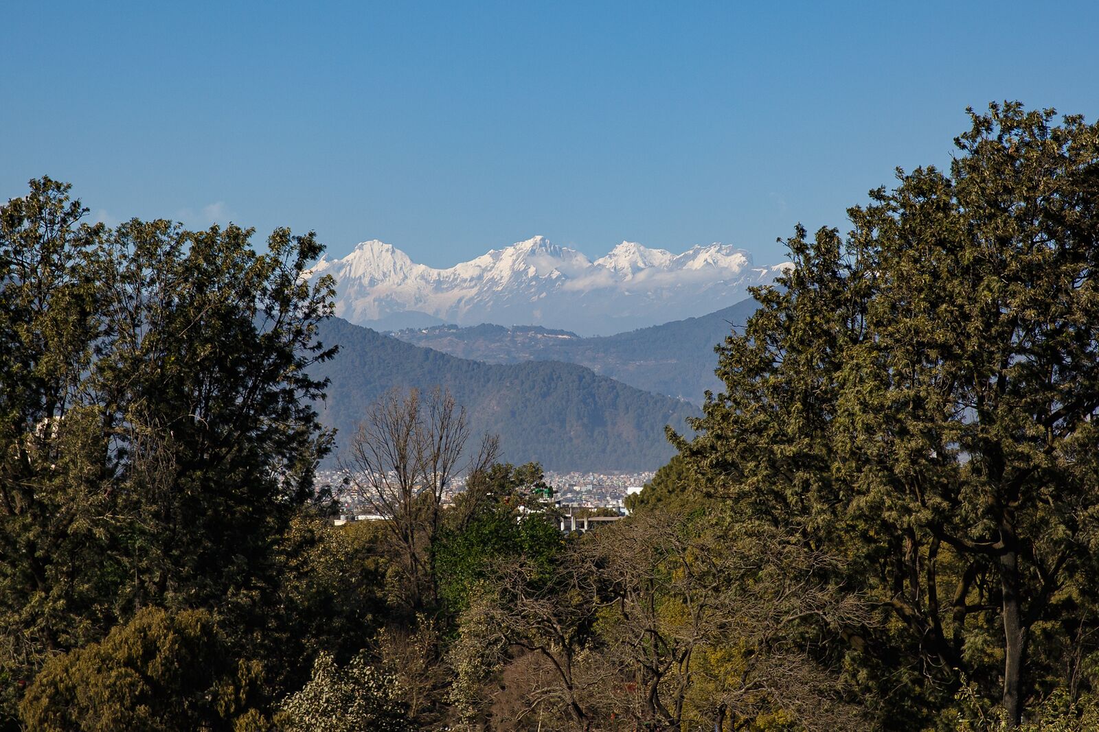 Paisagem montanhosa com picos nevados, áreas florestais densas e uma cidade ao longe.