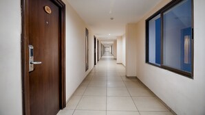 Um corredor comprido e estreito com uma porta de madeira identificada com o número 102 à esquerda e uma janela grande à direita.