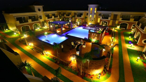 Área de piscina exterior bem iluminada, com bar e assentos, rodeada por um jardim ajardinado e um edifício com várias varandas.