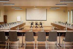 Uma sala de conferências com uma mesa comprida, várias cadeiras, garrafas de água, uma tela de projetor e um quadro branco.