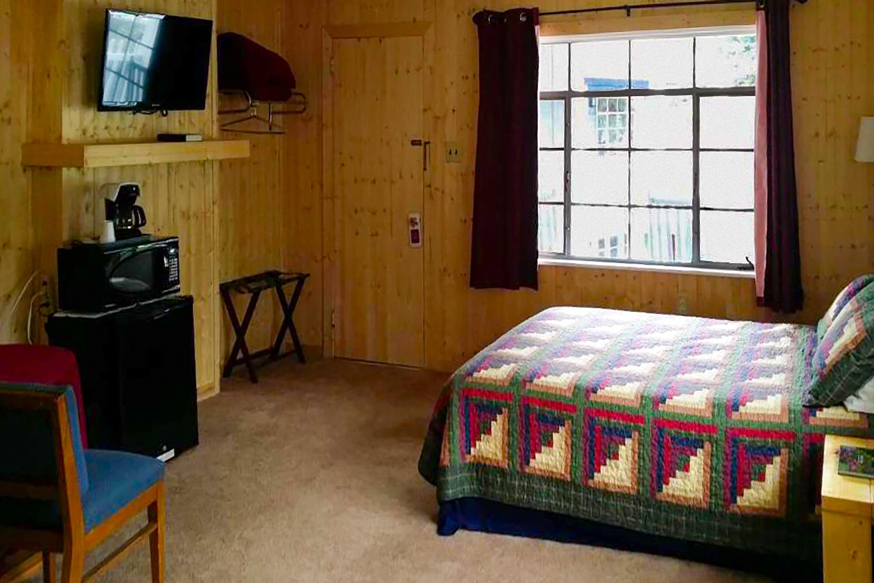 Quarto de madeira com cama, televisão, escrivaninha e cafeteira, e janela com cortinas.
