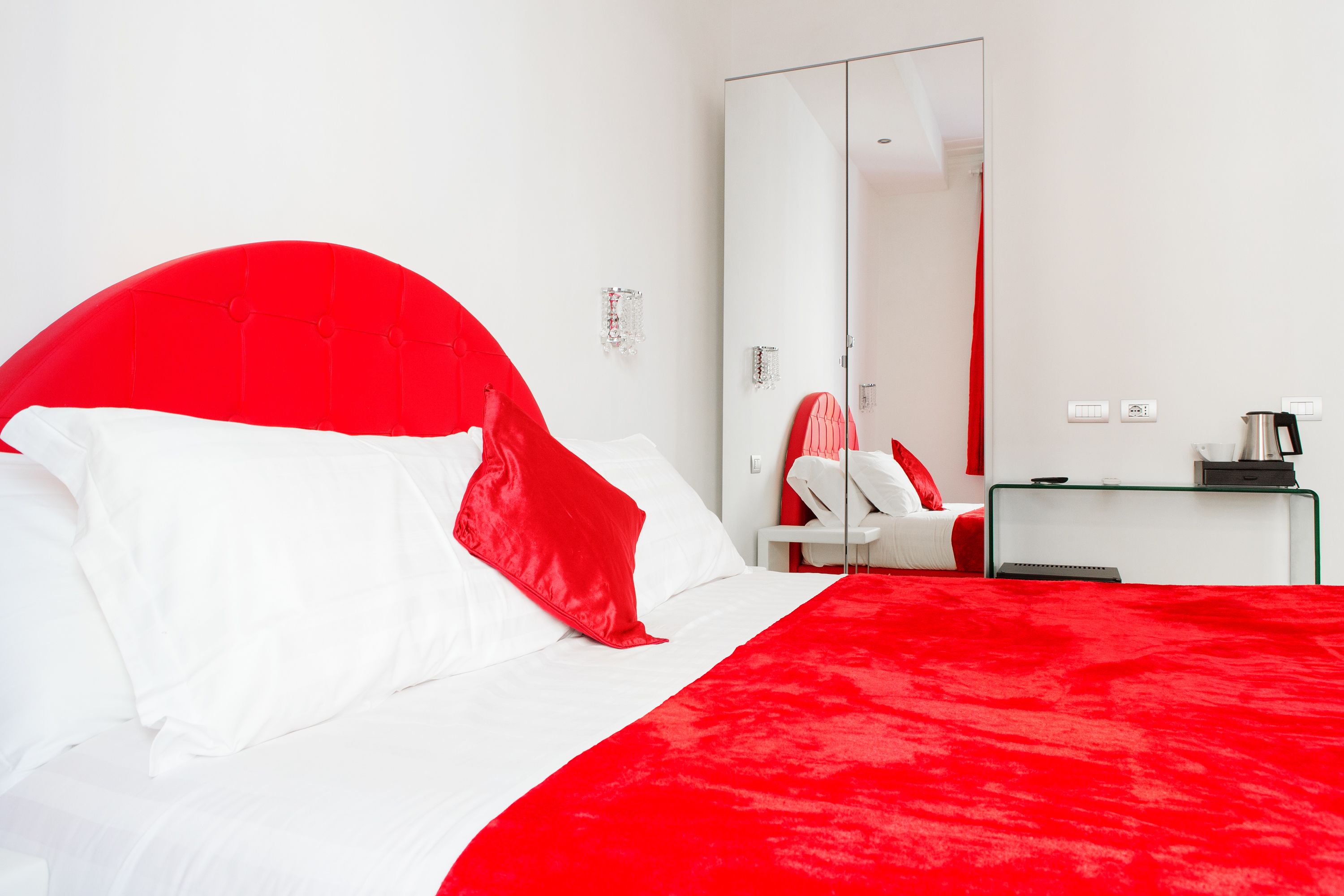 Quarto de hotel moderno com uma cama grande, decoração em tons de vermelho e branco, um guarda-roupa branco e uma mesa com cafeteira.