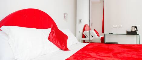 Quarto de hotel moderno com uma cama grande, decoração em tons de vermelho e branco, um guarda-roupa branco e uma mesa com cafeteira.