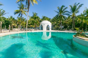 Uma piscina azul cristalina com uma estrutura em forma de cúpula no centro, rodeada por palmeiras.
