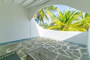 Quarto com piso de pedra, um banco de madeira e vista para palmeiras através de uma janela curva.