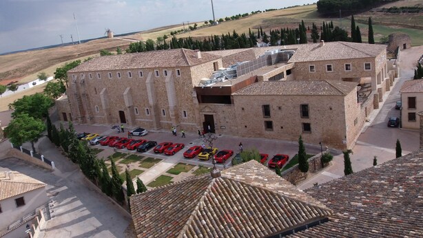 Um edifício grande com vários andares, um estacionamento com diversos carros e uma paisagem montanhosa ao fundo.