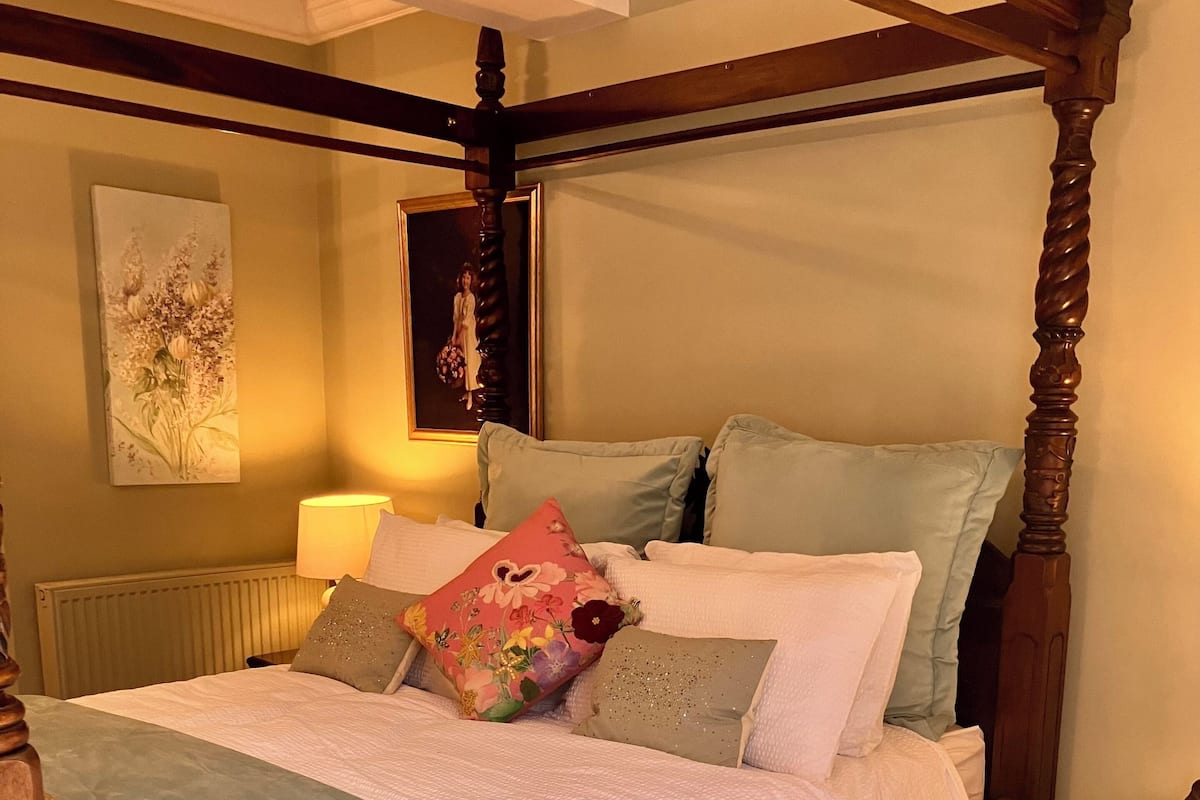 Uma cama de dossel com cabeceira de madeira, mesa de cabeceira, abajur e um quadro na parede.