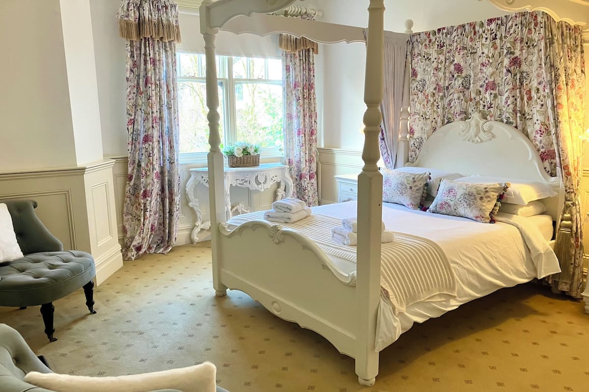 Uma cama de dossel com cortinas florais, uma poltrona cinza e uma mesinha com um cesto.