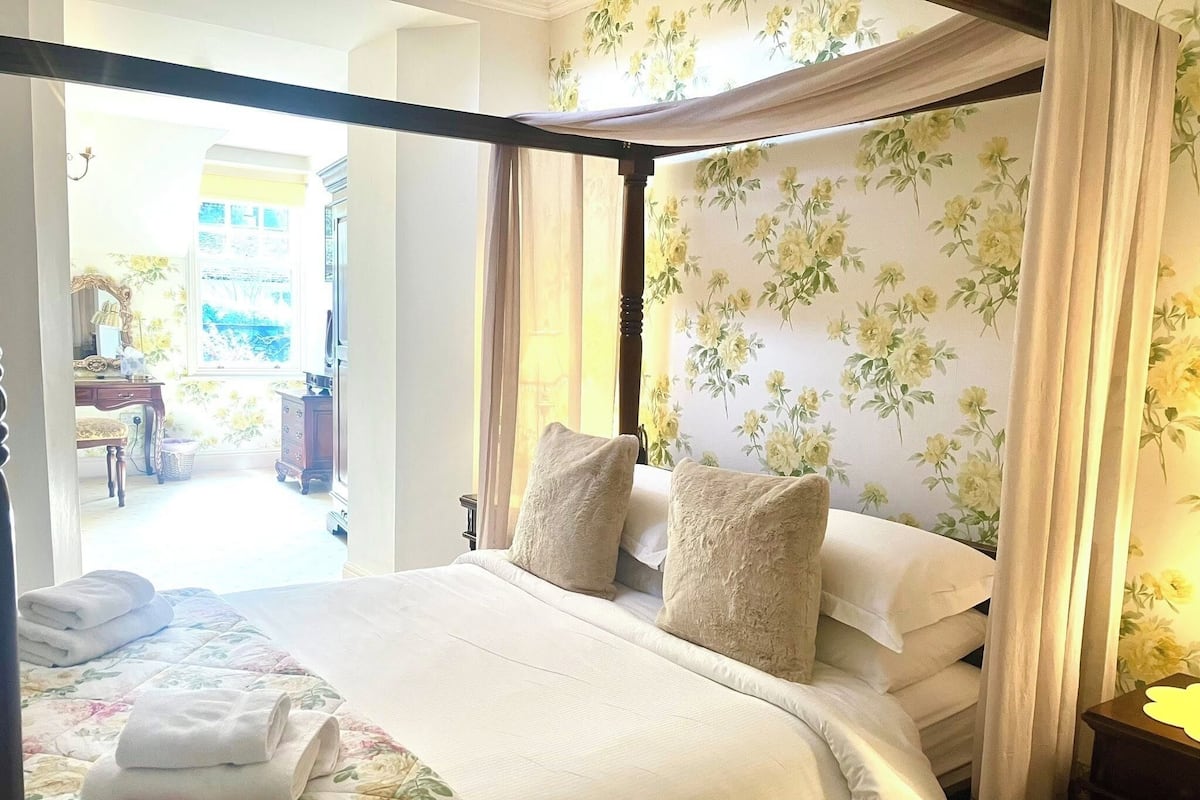 Uma cama com dossel, papel de parede floral, uma mesa de cabeceira e vista do interior do quarto.