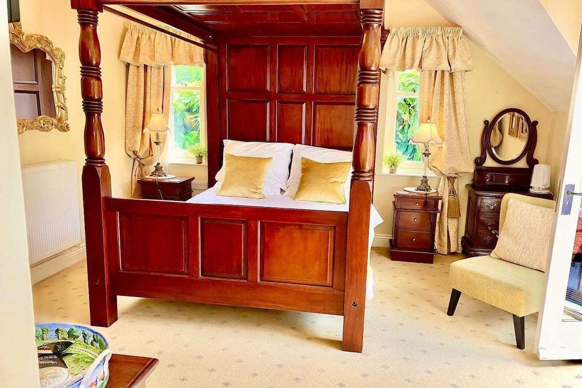 Uma cama com dossel e estrutura de madeira, uma mesa de cabeceira com abajur, uma cadeira e um penteado com espelho.