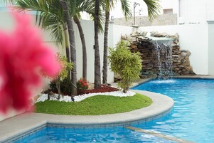Piscina com uma cascata de pedra, rodeada por palmeiras e um pequeno jardim.