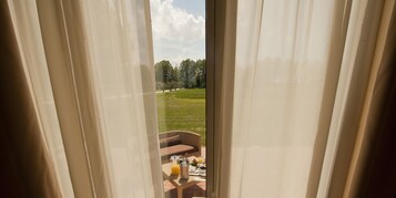 Varanda com mesa posta para o pequeno-almoço, uma cadeira de vime e vista para um campo relvado e árvores.