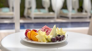 Uma bandeja de frutas sobre uma mesa, com uma piscina e cadeiras de descanso ao fundo.