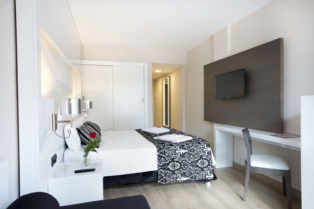 Quarto de hotel moderno com cama, mesa de cabeceira, cadeira, escrivaninha e televisão.