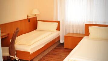 Quarto de hotel com duas camas de solteiro, cabeceira de madeira, escrivaninha com cadeira e janela com cortinas leves.