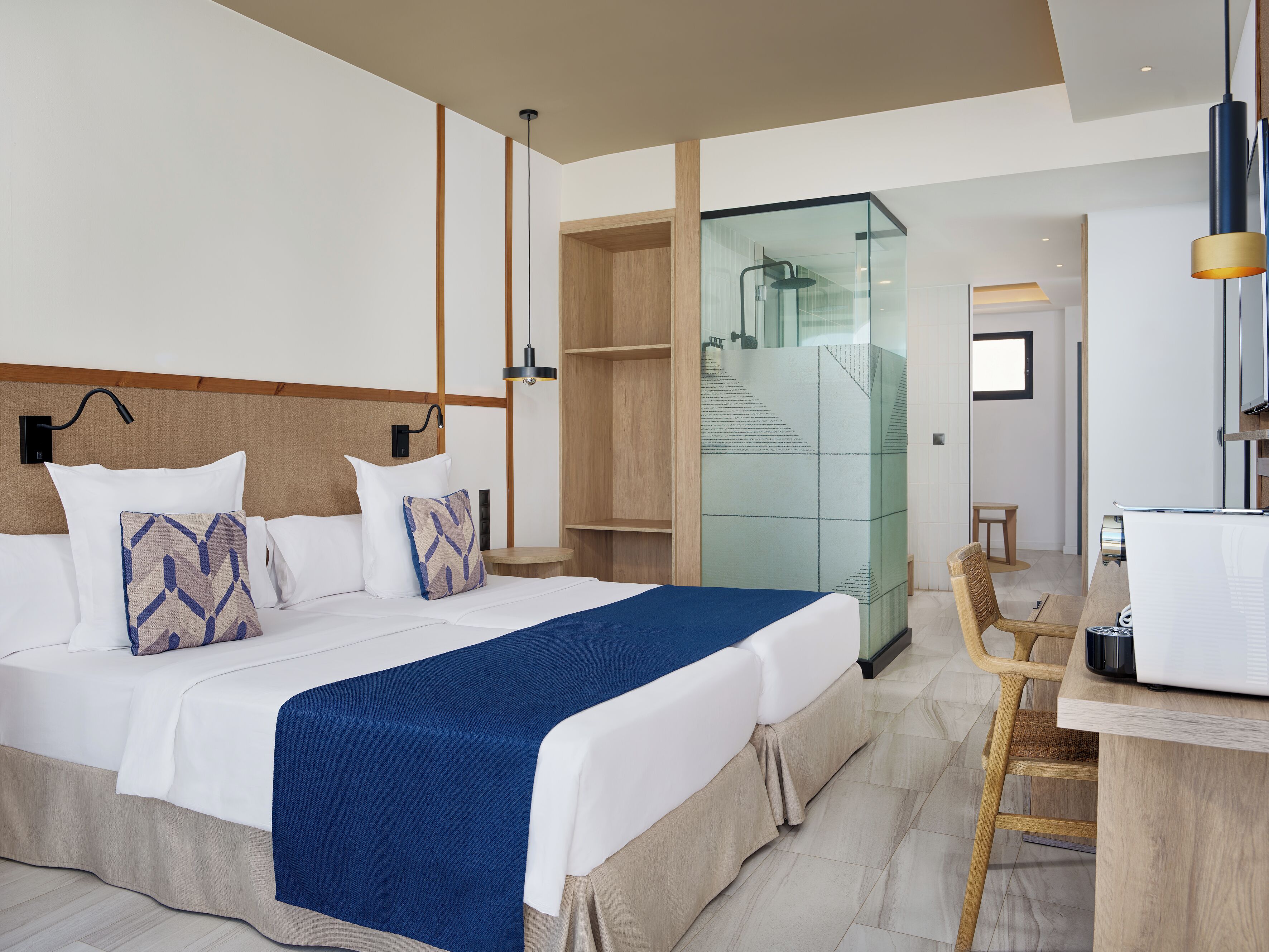 Quarto de hotel moderno com uma cama grande, chuveiro de vidro, escrivaninha de madeira e uma cadeira.