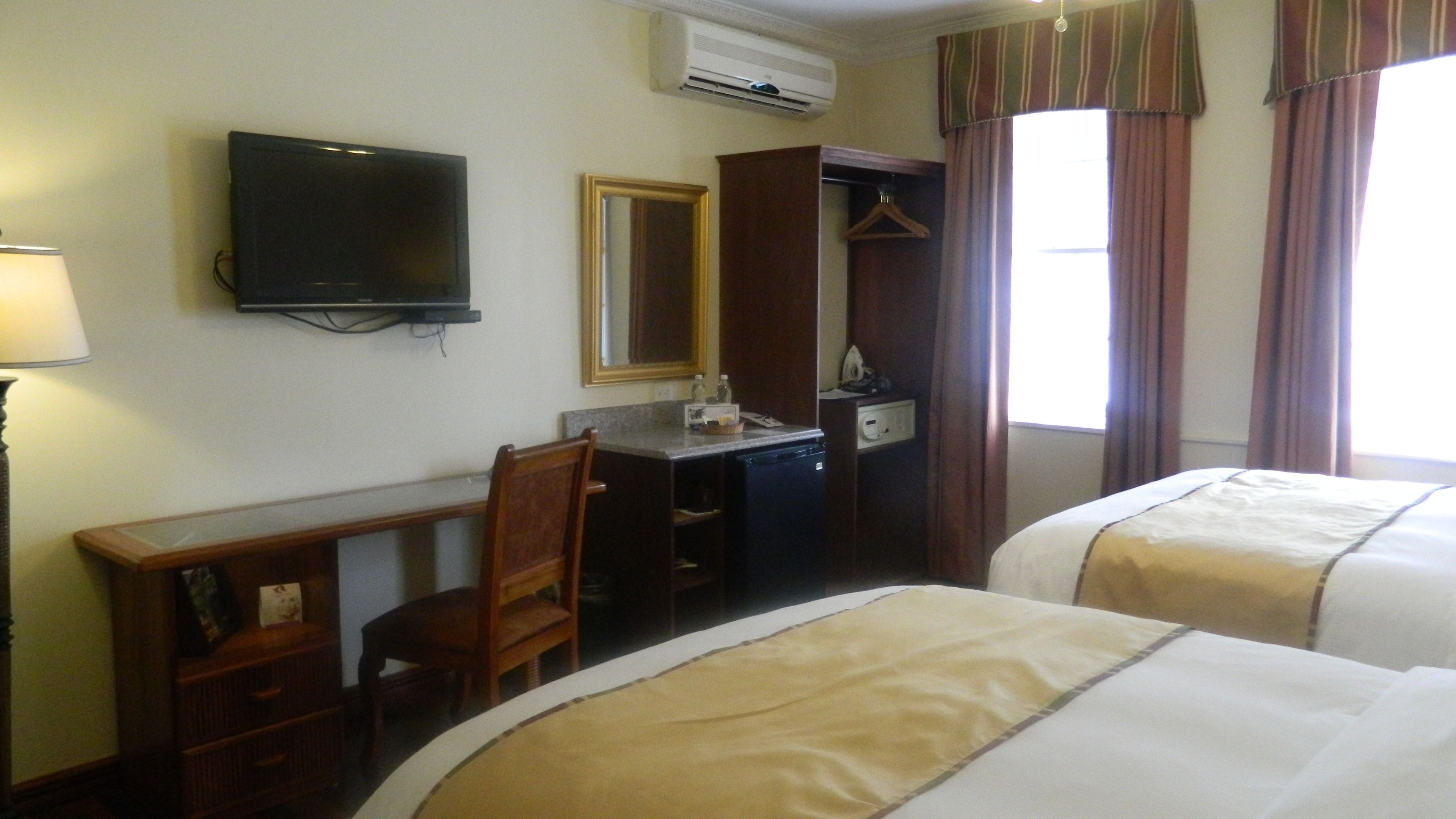 Quarto de hotel com duas camas, uma escrivaninha com cadeira, televisão, espelho, guarda-roupa e janela com cortinas.