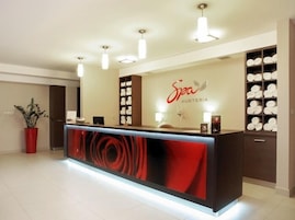 Uma área de recepção com design moderno, destacando um balcão central com um design abstrato vermelho, uma placa com a inscrição "Spa Austria" e prateleiras com toalhas dobradas.