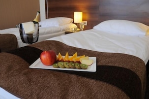 Quarto de hotel com cama, mesa de cabeceira com uma garrafa de champanhe e um prato de frutas.