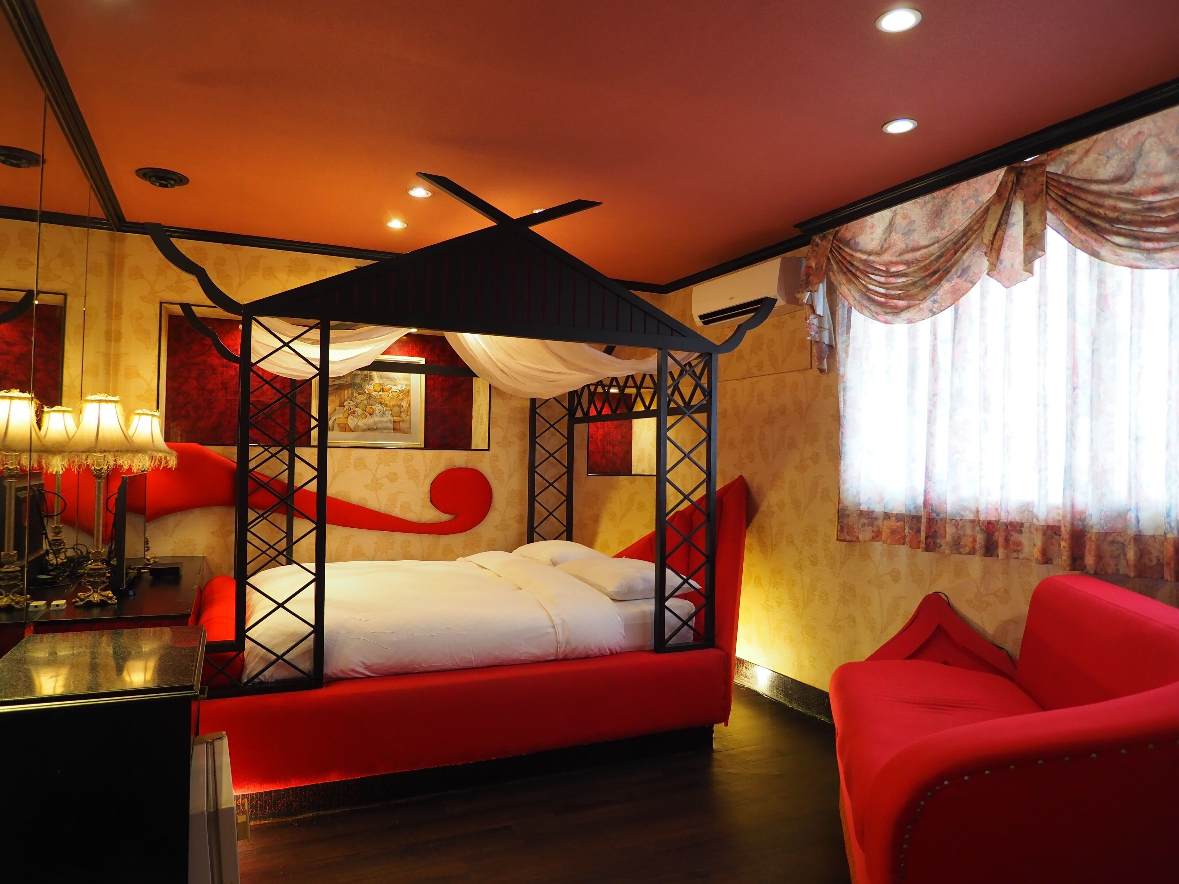 Uma cama de dossel com cortina, um sofá vermelho e uma escrivaninha com luminárias.