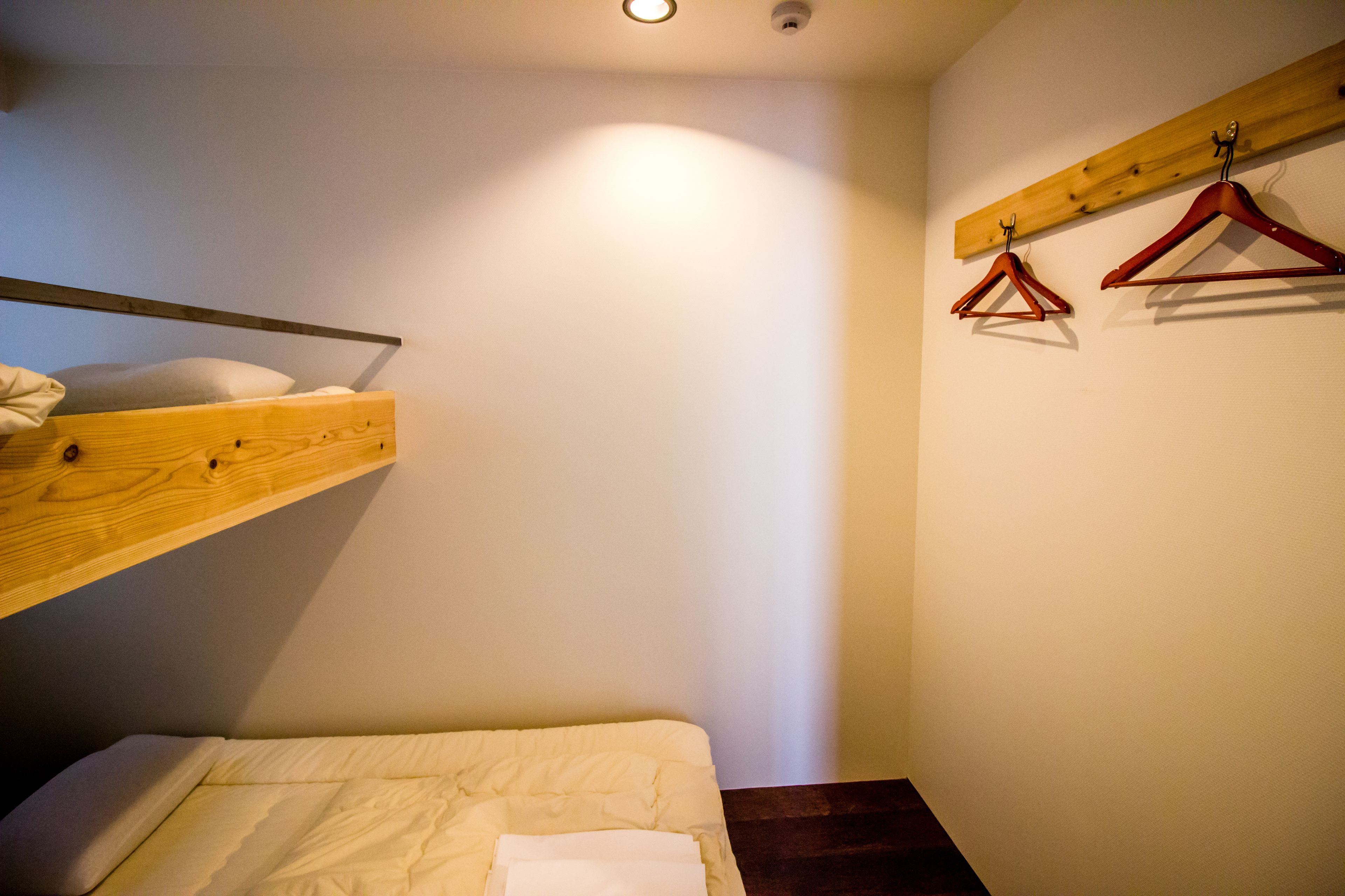Quarto pequeno e bem iluminado com uma cama beliche, uma prateleira de madeira com cabides e uma parede simples.