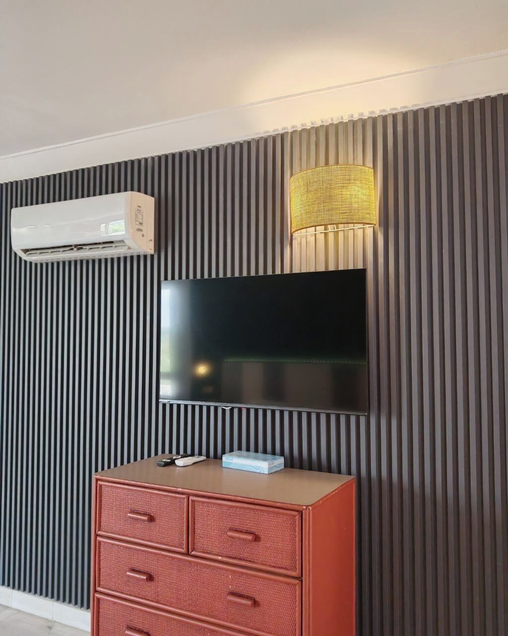Quarto moderno com televisão de tela plana fixada em uma parede com painéis verticais, um armário vermelho com gavetas e um aparelho de ar condicionado.