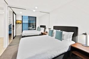 Quarto de hotel moderno com duas camas, um espelho grande, televisão e vista para o mar.
