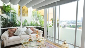 Uma sala de estar ampla com sofá branco, mesa de centro de vidro e vista para a paisagem urbana através de grandes janelas.