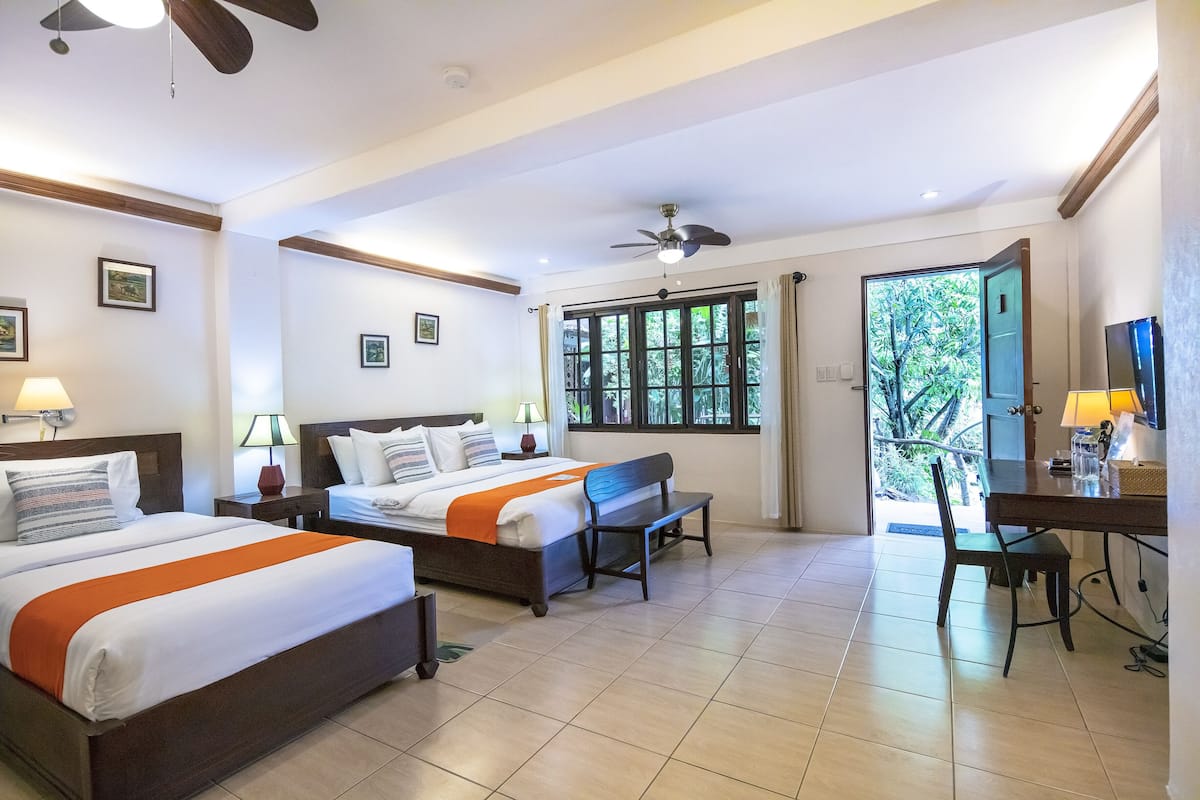 Quarto de hotel com duas camas, uma escrivaninha, uma cadeira, ventilador de teto e uma grande janela com vista para o verde.