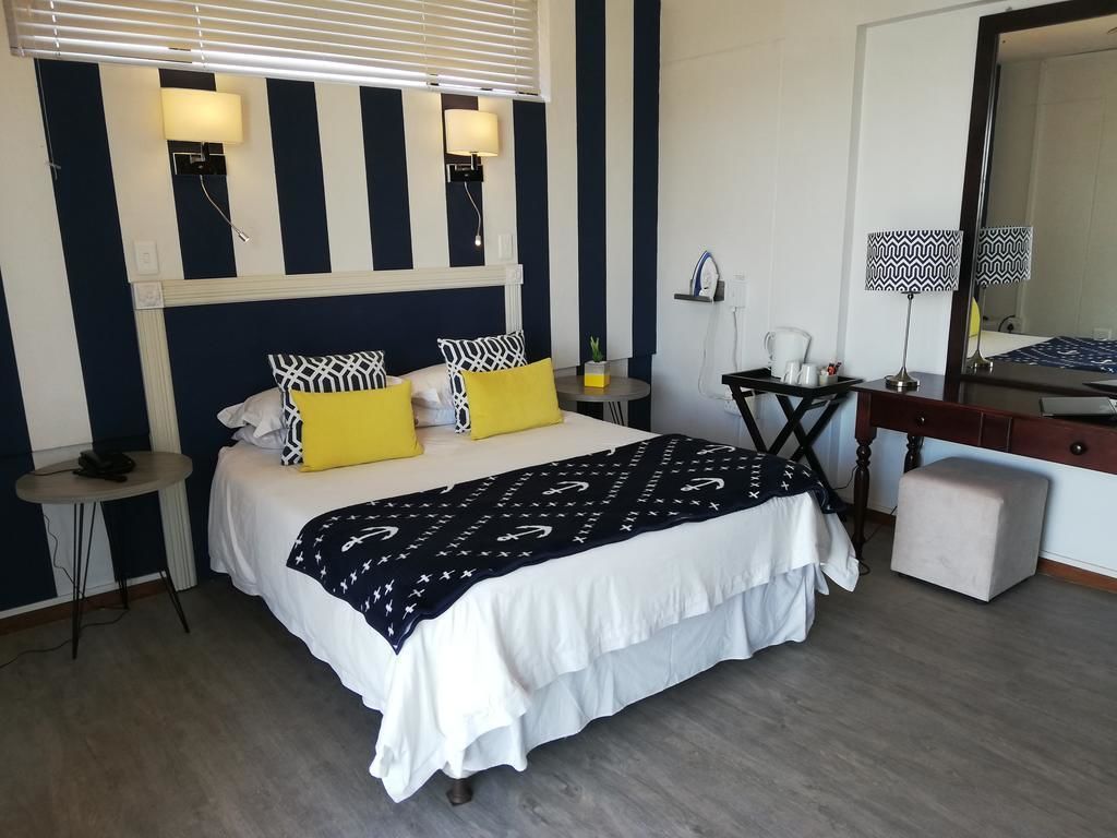 Quarto de hotel com cama, mesas de cabeceira, escrivaninha, cadeira, espelho e um banquinho.