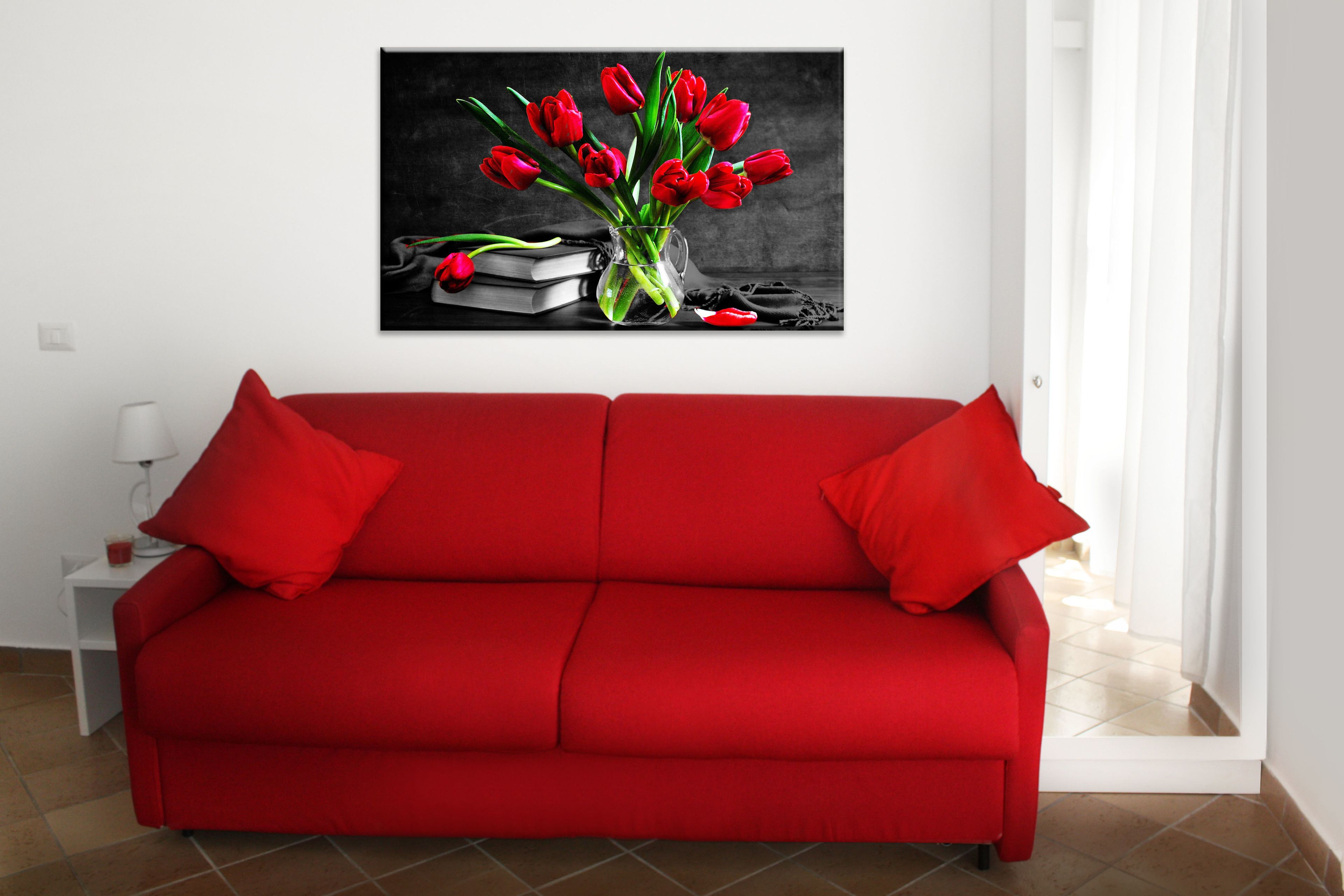 Um sofá vermelho com almofadas combinando, uma pintura em preto e branco de tulipas vermelhas e uma mesinha de cabeceira branca com uma luminária.