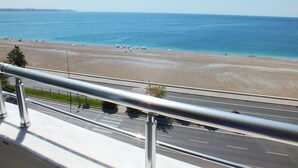 Vista da praia a partir de uma varanda com guarda-corpo, uma estrada em primeiro plano e céu claro.