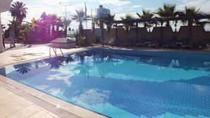 Piscina com espreguiçadeiras e palmeiras, cercada por um deck ladrilhado e uma parede de pedra.